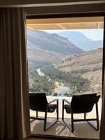 Quarto com uma grande janela com vista para uma paisagem montanhosa. Há duas cadeiras pretas e uma mesinha em frente à janela.