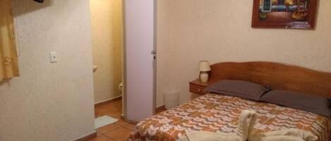 Quarto com cama, mesa de cabeceira, abajur, televisão fixada na parede e porta que dá para outro cômodo.