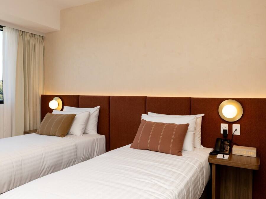 Quarto de hotel com duas camas, cada uma com lençóis brancos e almofadas castanhas. Há luminárias de cabeceira com luzes redondas, uma mesa de cabeceira de madeira com telefone e relógio digital, e uma janela com cortinas.