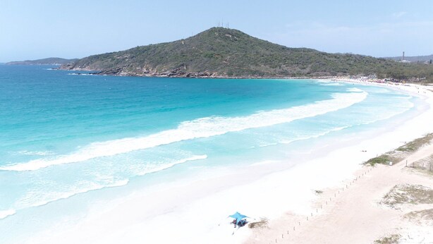 Uma praia com águas turquesa cristalinas, uma faixa de areia e uma colina com vegetação e construções ao fundo.