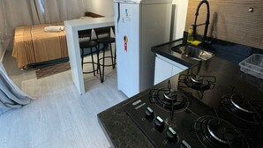 Uma cozinha compacta com geladeira, fogão e micro-ondas. Há uma pequena área de jantar com mesa e cadeiras. O quarto possui uma janela com cortinas.