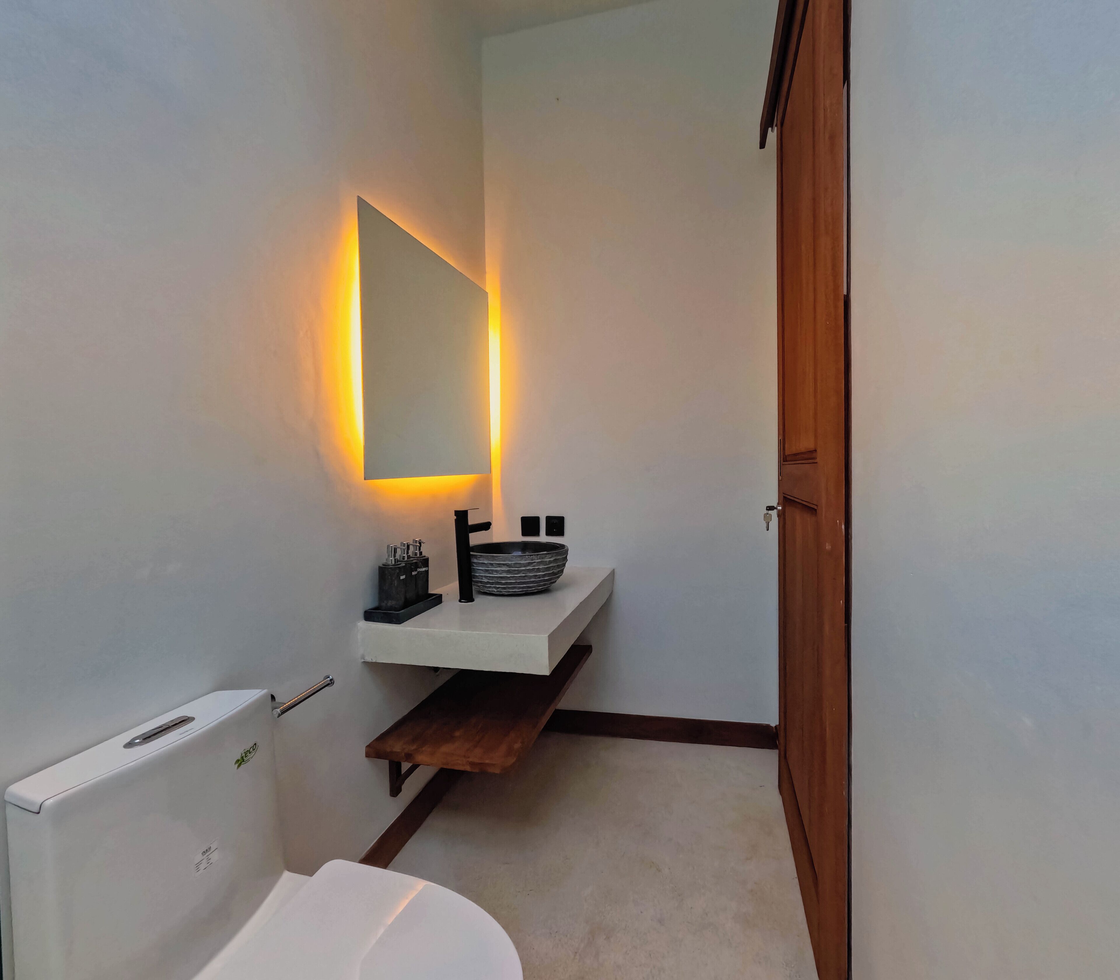 Um banheiro moderno com pia branca, espelho com iluminação embutida, vaso sanitário e um banco de madeira.
