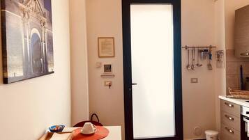 Um corredor estreito com uma porta que dá para uma área de cozinha. Há uma moldura com um quadro na parede, uma pequena mesa com uma xícara e uma bandeja de petiscos, e uma lixeira perto da porta.