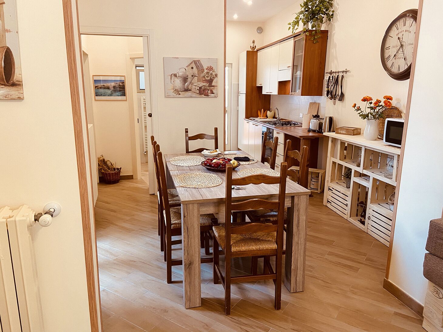Uma sala de jantar com mesa e cadeiras de madeira, uma cozinha com armários de madeira e um relógio, e um corredor que dá para outro quarto.