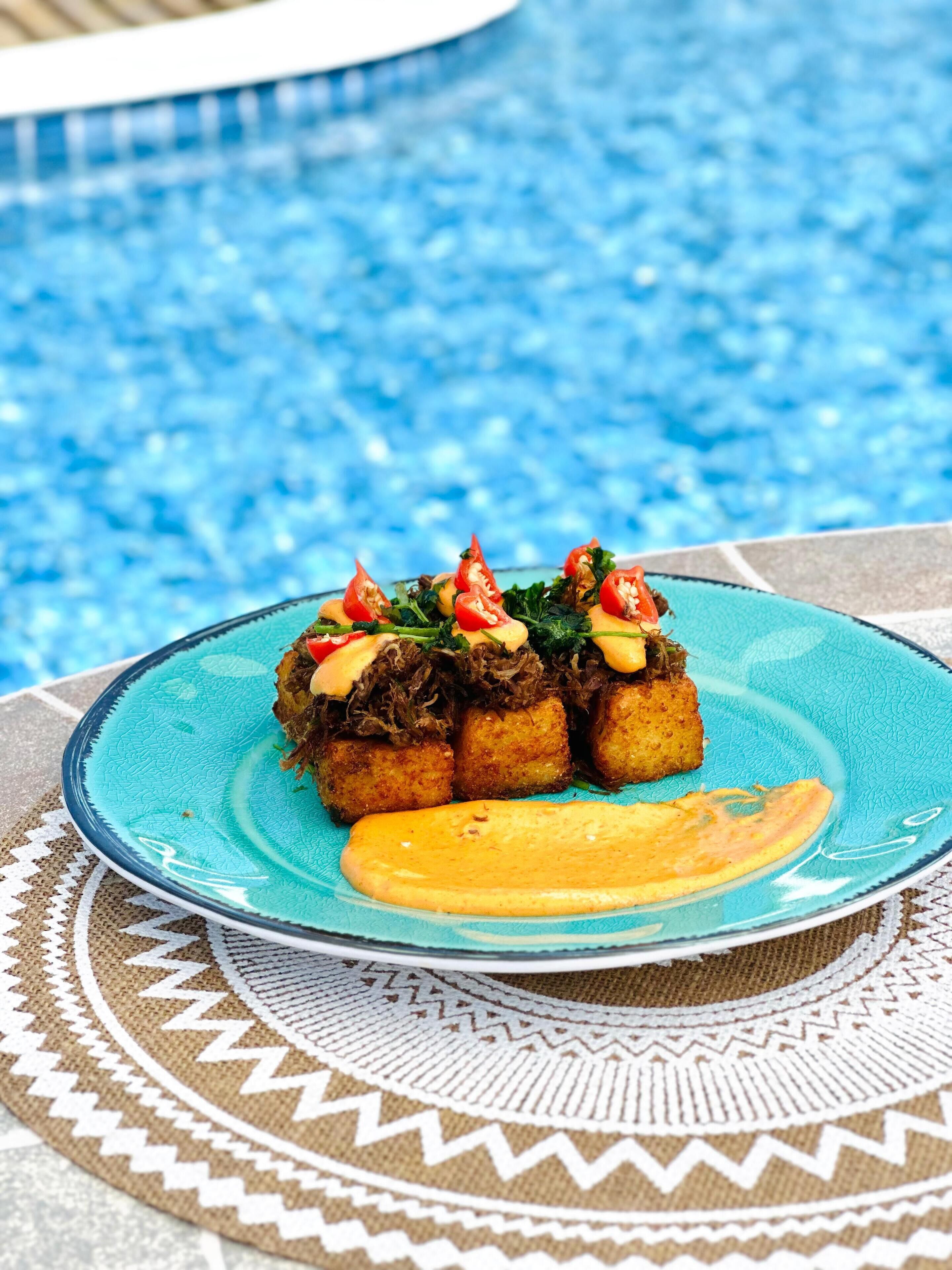 Prato de comida com borda azul, apoiado em um jogo de mesa estampado junto a uma piscina.
