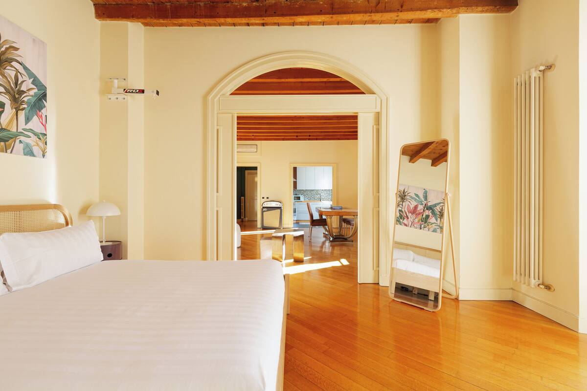 Quarto de hotel com cama, piso de madeira, escrivaninha com cadeira, radiador e um quadro na parede.