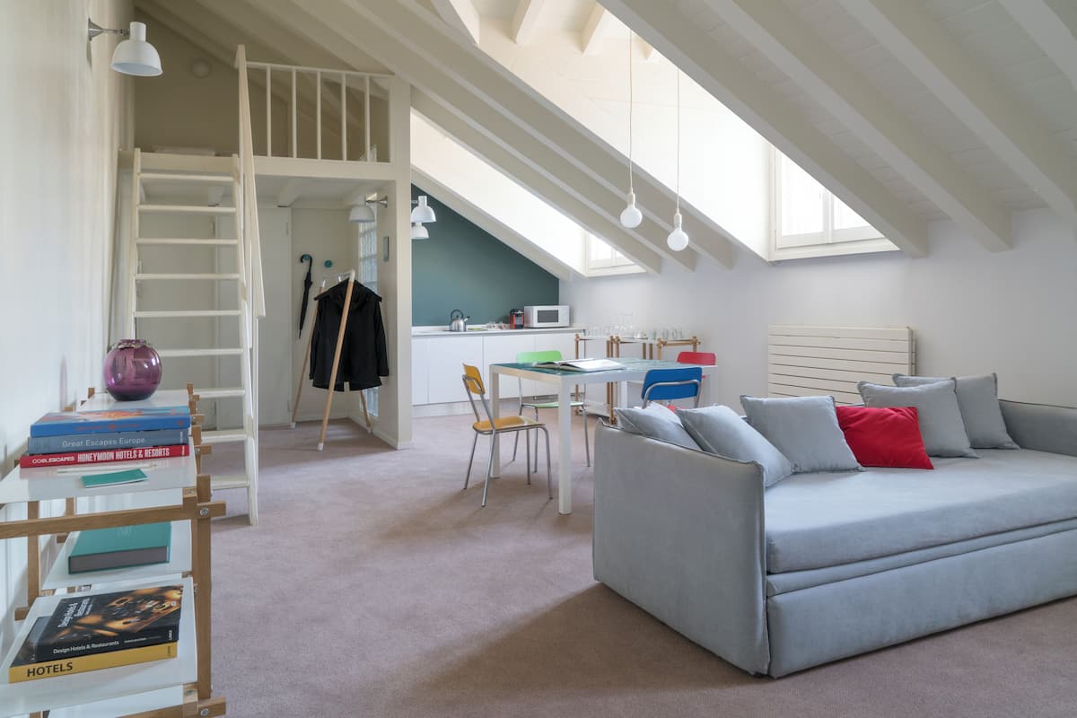 Quarto estilo loft com sofá cinza, uma mesa de jantar e escada para um andar superior.