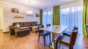 Uma sala de estar moderna com um sofá, uma mesa de jantar e cadeiras. Há objetos decorativos nas paredes e uma janela grande com cortinas.