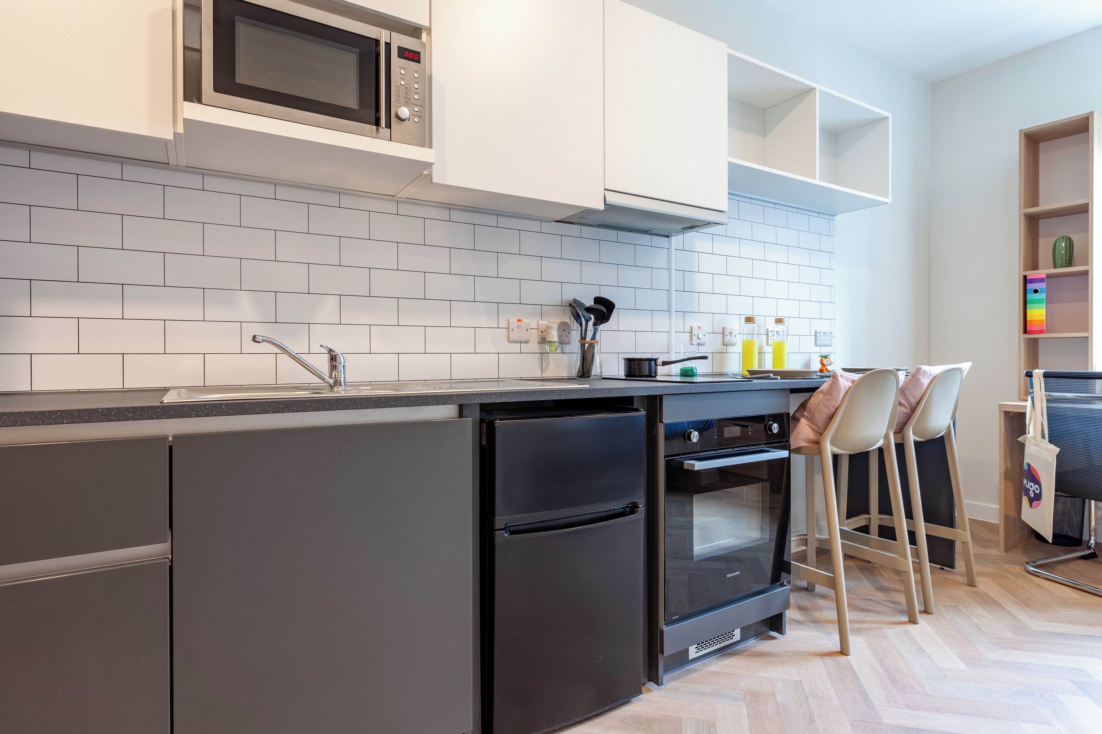Cozinha moderna com micro-ondas, forno e frigorífico. Há duas cadeiras altas de bar e uma pequena estante com objetos decorativos.