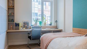 Quarto com cama, escrivaninha, cadeira e janela com vista para edifícios.