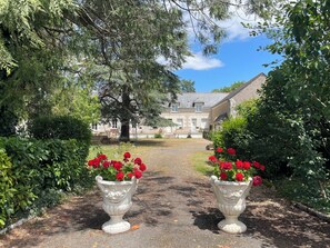 Uma trilha leva a um edifício com entrada para um jardim, com dois grandes vasos de flores vermelhas.
