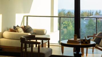 Uma sala de estar moderna com um sofá, duas cadeiras de madeira, uma mesinha redonda com livros e uma varanda com vista para o verde e o mar.