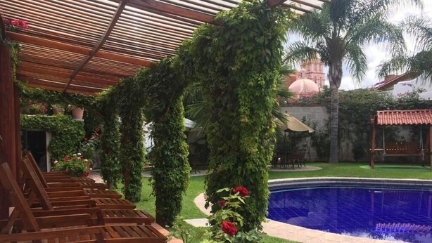 Uma área de varanda coberta com cadeiras de lounge de madeira, piscina e vegetação exuberante.