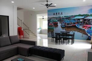 Sala de estar com sofá, puf e área de jantar com cadeiras. Uma escada é visível ao fundo.