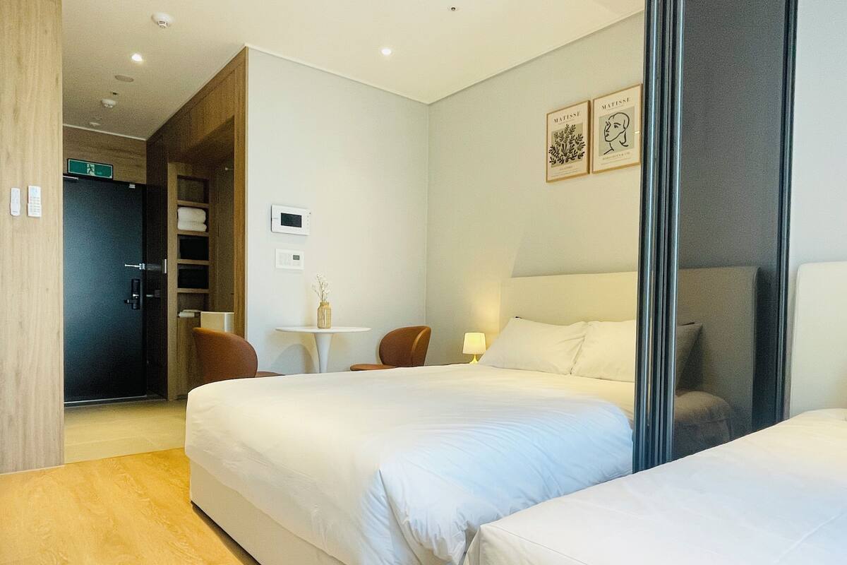 Quarto de hotel com duas camas, uma mesinha com um vaso e uma cadeira.