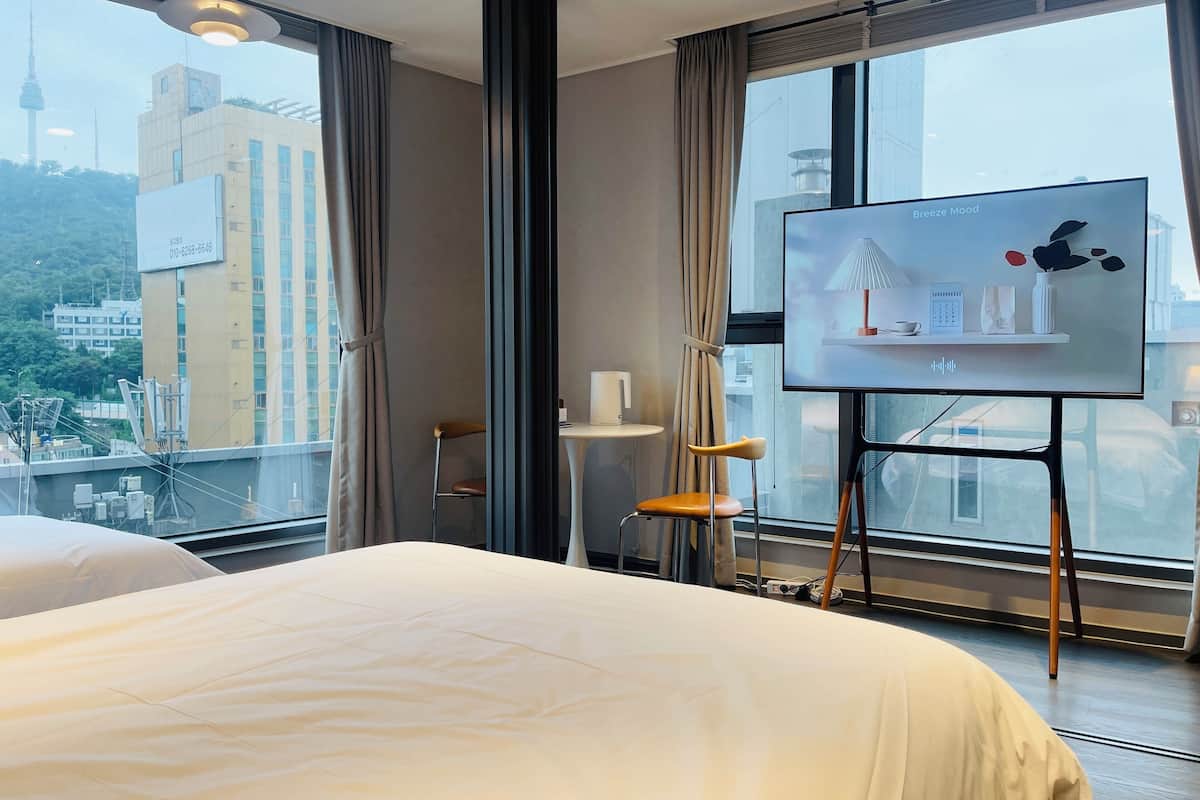 Quarto de hotel com uma cama grande, uma escrivaninha com cadeira, uma TV sobre suporte e uma janela com vista para a cidade.