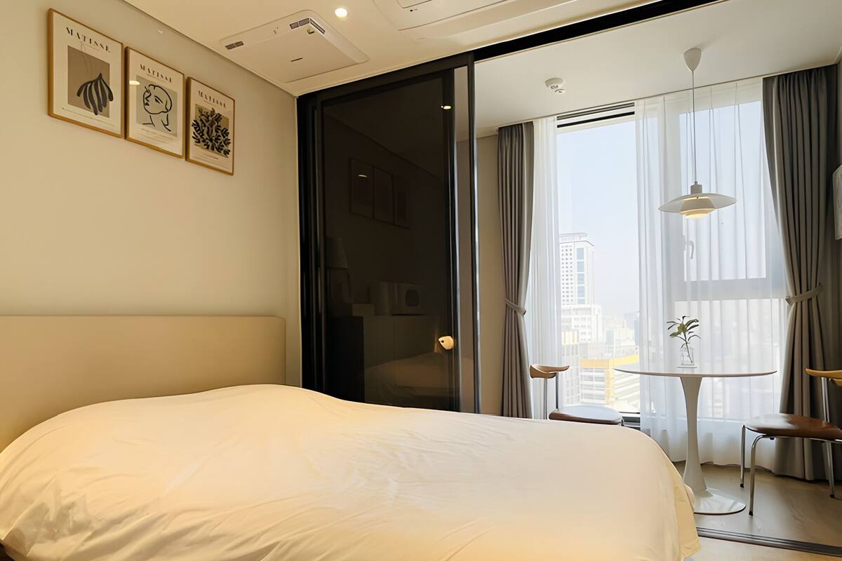 Quarto de hotel com uma cama grande, uma porta de vidro deslizante para uma varanda, uma pequena mesa com uma cadeira e arte emoldurada na parede.
