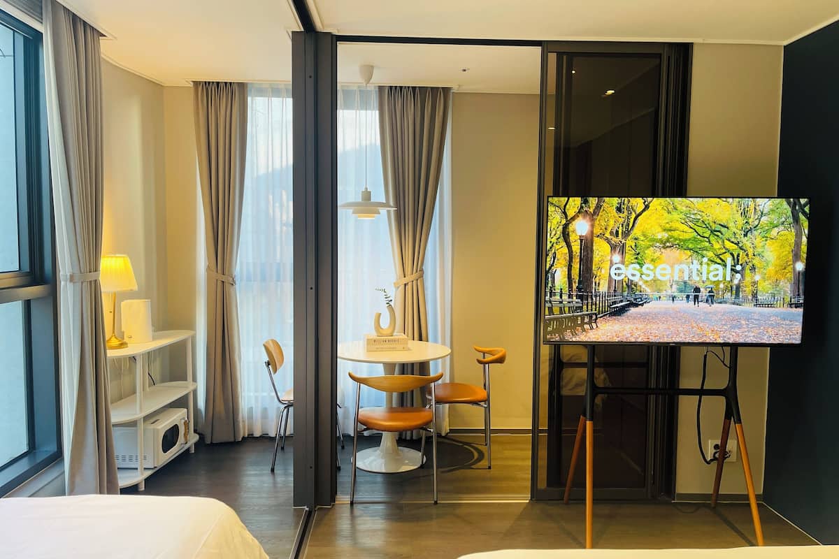 Quarto de hotel com cama, televisão exibindo uma cena de parque, área de refeições com mesa e cadeiras e micro-ondas em uma prateleira.