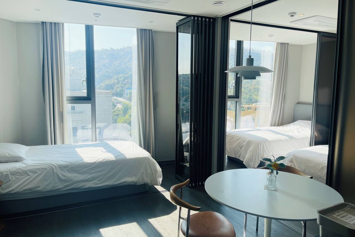 Quarto de hotel moderno com duas camas, uma mesa redonda e vista para a paisagem urbana.