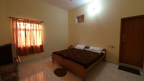 Quarto com cama, ventoinha no teto, janela com cortinas, porta e pequena mesa com cadeira.