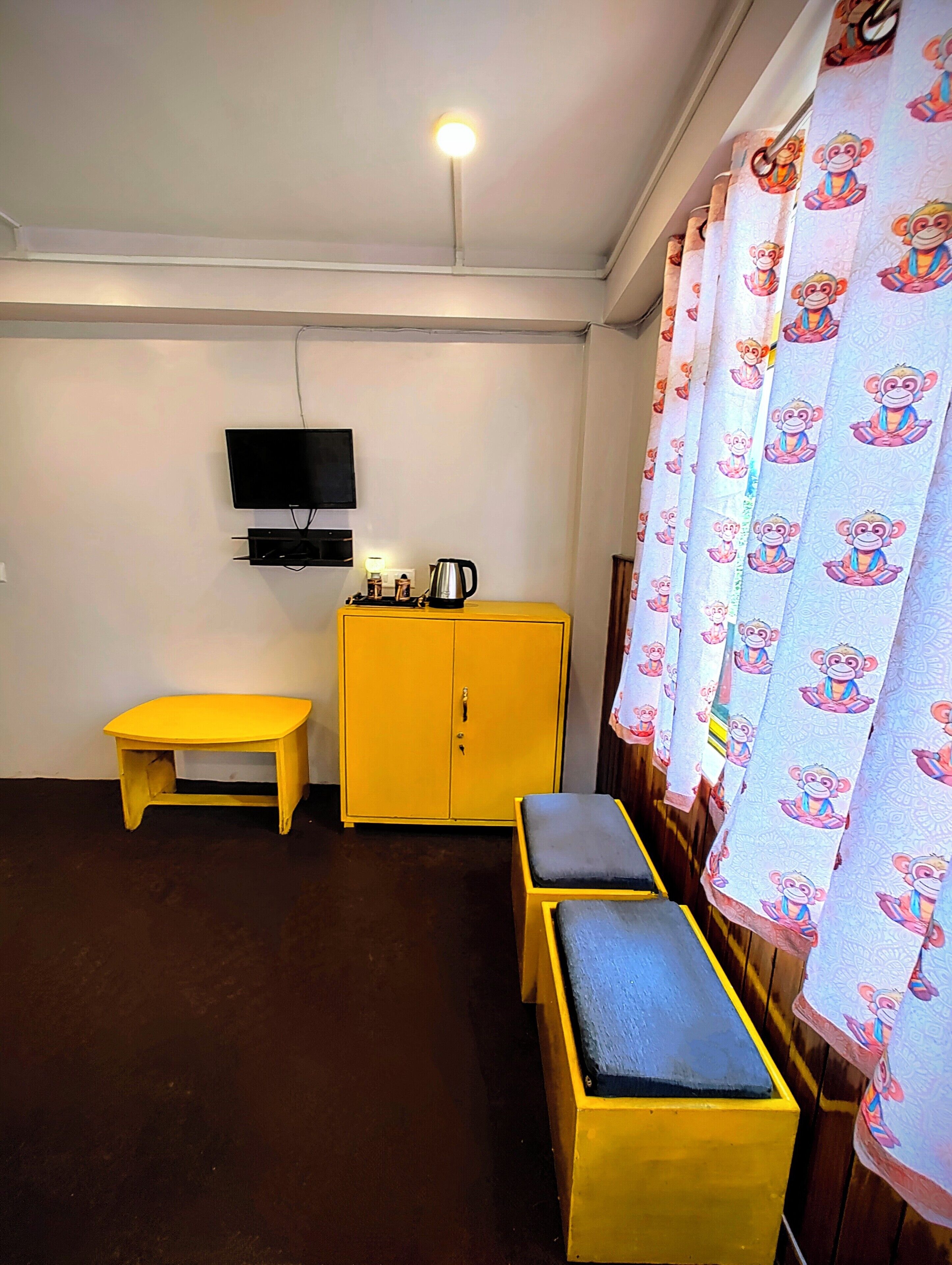 Um quarto com um armário amarelo, um banco, uma televisão fixada na parede e cortinas com padrão de macaco.