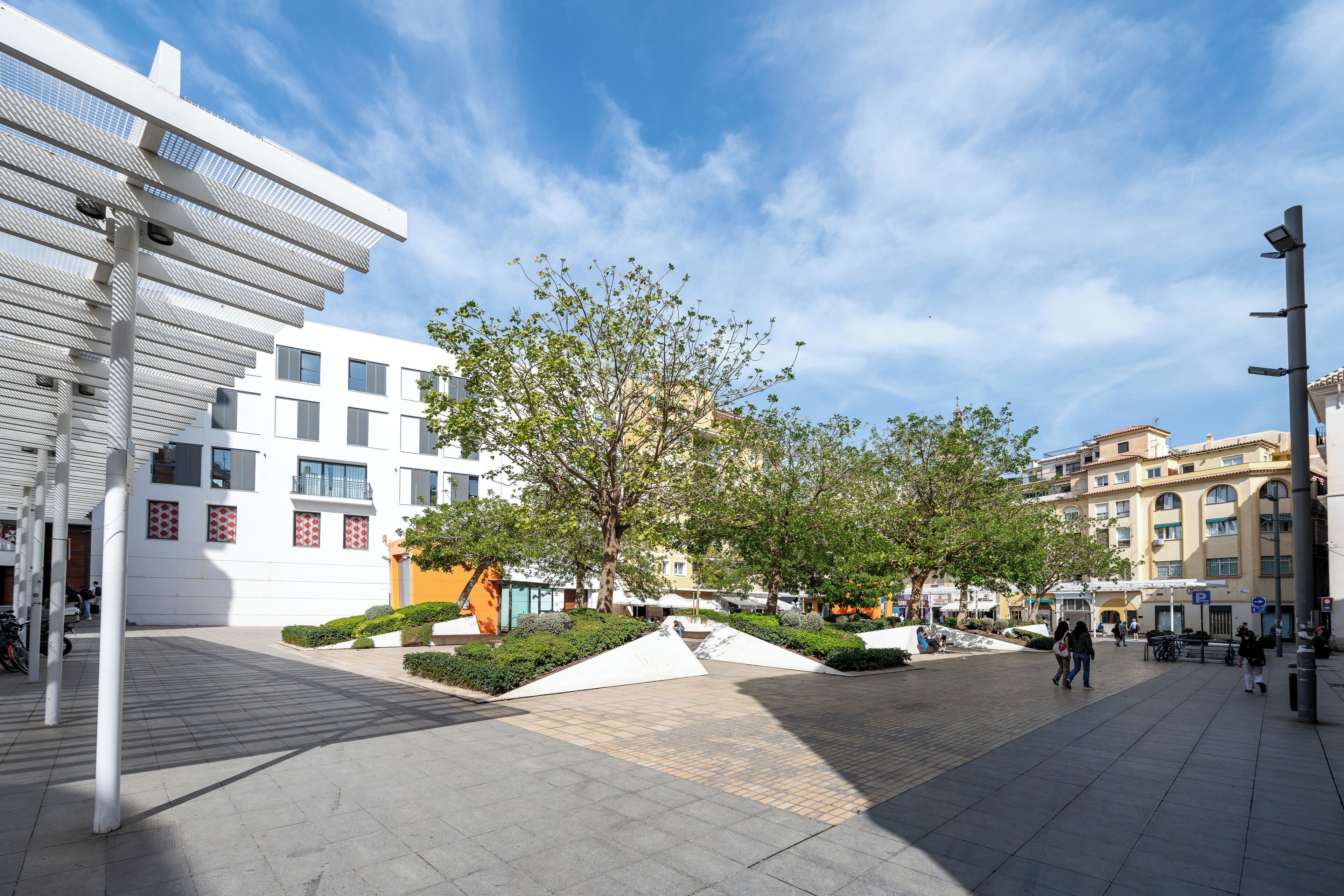 Uma ampla praça urbana com arquitetura moderna, uma área central arborizada e céu claro.
