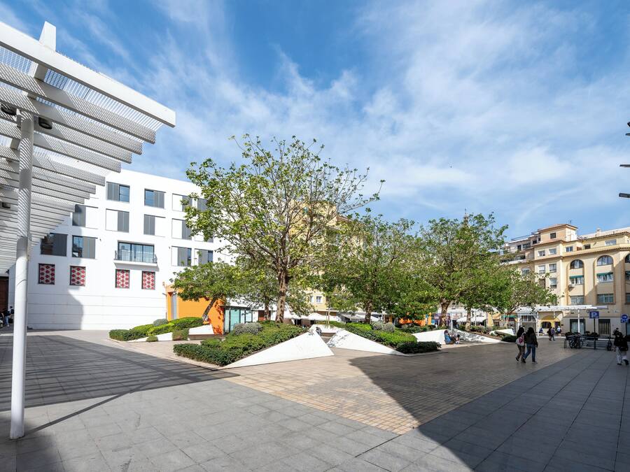 Uma ampla praça urbana com arquitetura moderna, uma área central arborizada e céu claro.