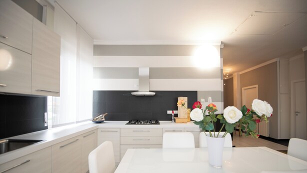 Uma cozinha moderna com armários brancos, painel de fundo preto e uma área de refeições com mesa e cadeiras brancas.