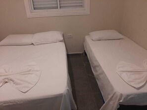 Duas camas de solteiro com roupa de cama branca, uma janela acima e piso de azulejos.