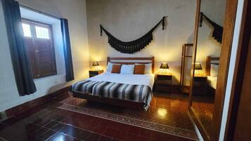 Quarto de hotel com uma cama grande, duas mesas de cabeceira com luminárias, uma janela com cortinas e piso ladrilhado.