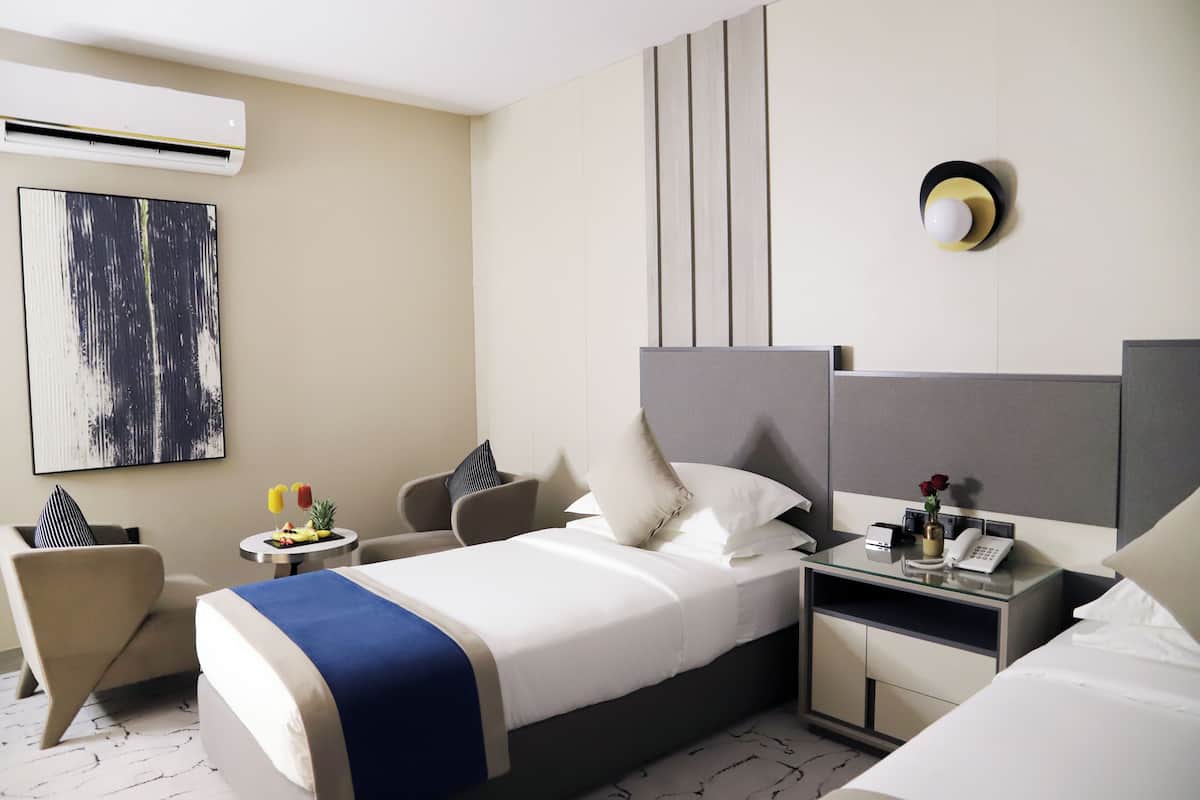 Quarto de hotel moderno com uma cama grande, uma escrivaninha com telefone, uma cadeira e uma pequena mesa com frutas e um vaso de flores.