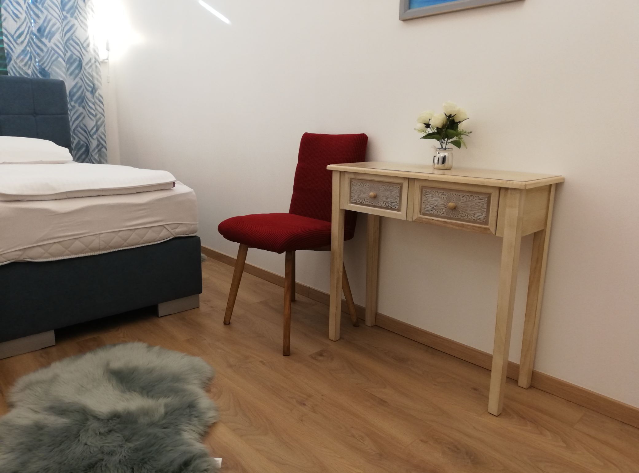 Um quarto com uma cama, uma cadeira vermelha, uma escrivaninha de madeira com duas gavetas e um pequeno vaso com flores na escrivaninha.