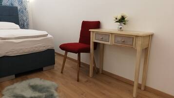 Um quarto com uma cama, uma cadeira vermelha, uma escrivaninha de madeira com duas gavetas e um pequeno vaso com flores na escrivaninha.