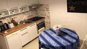 Uma cozinha compacta com armários brancos, bancada de madeira, pia e fogão. Há uma mesa com toalha azul e um vaso com flores. Um letreiro na parede diz 'HELLO Summer'.