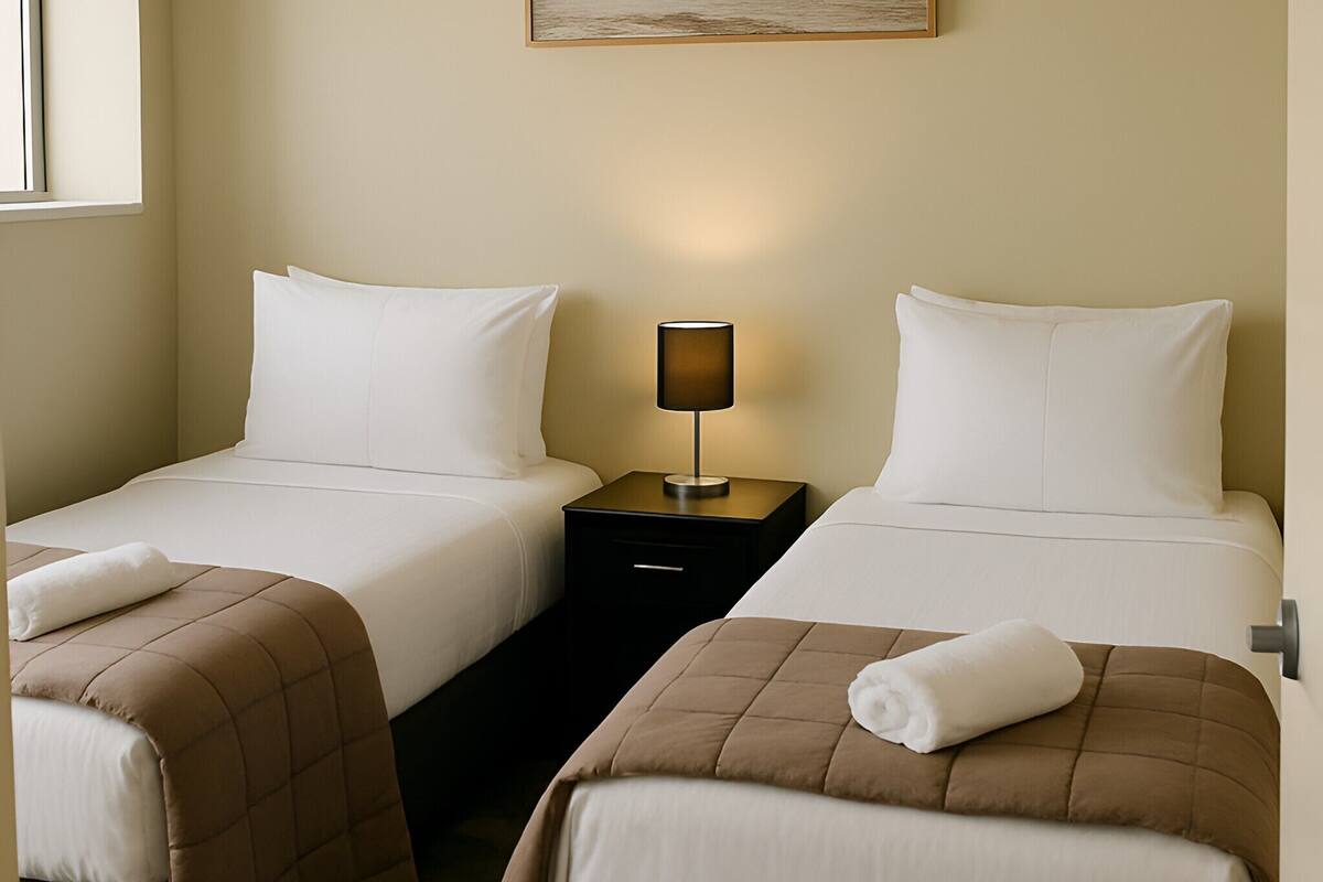 Quarto de hotel com duas camas, uma mesinha de cabeceira com abajur e um quadro na parede.