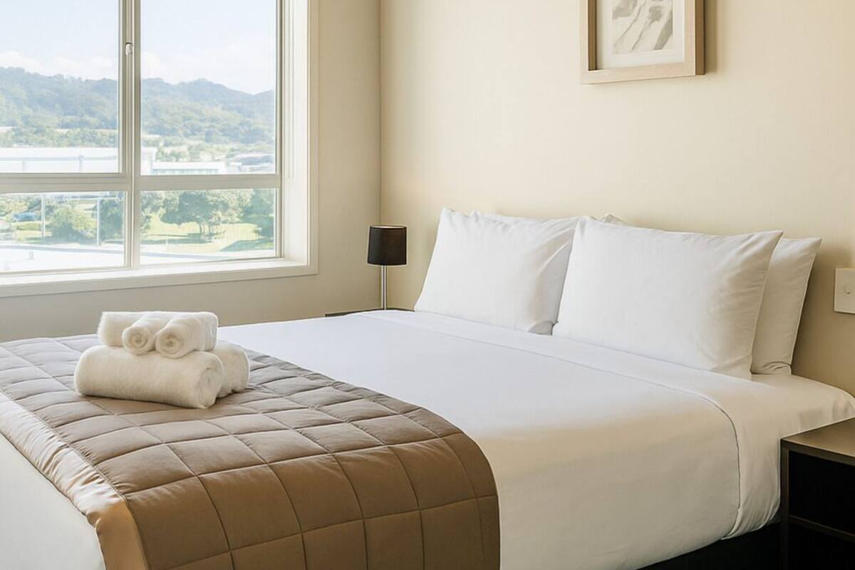 Cama bem arrumada com colcha marrom acolchoada e almofadas brancas, um par de toalhas enroladas na cama, um abajur e um quadro na parede.