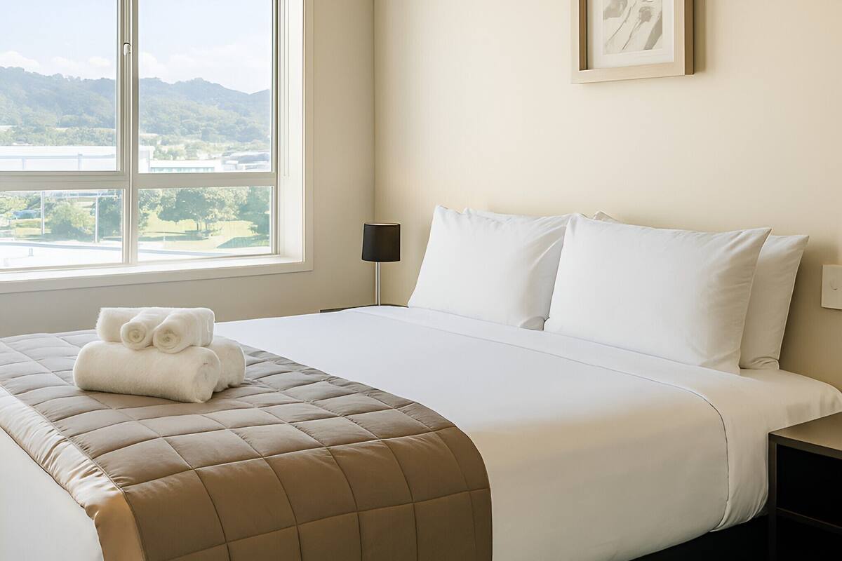 Quarto de hotel com cama, janela com vista para vegetação e montanhas, abajur e quadro na parede.