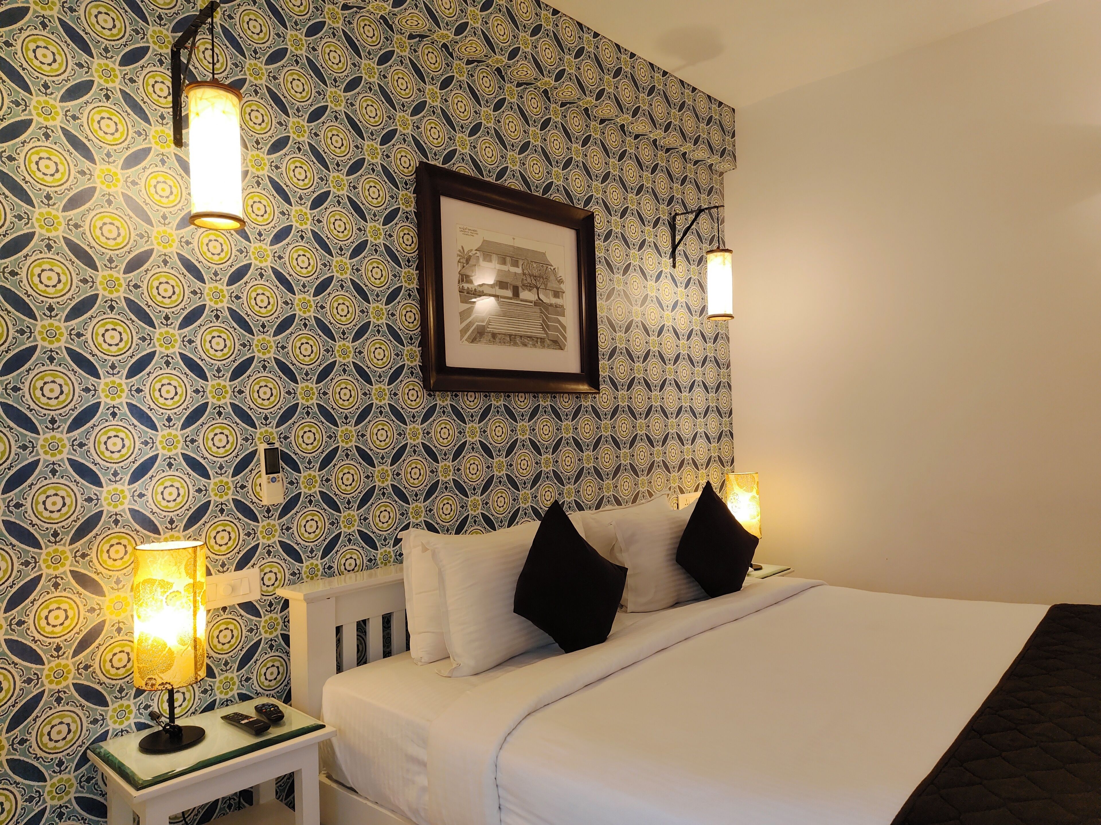 Quarto de hotel com papel de parede estampado, um quadro na parede, uma cama com roupa de cama branca e almofadas pretas, e duas luminárias de cabeceira.