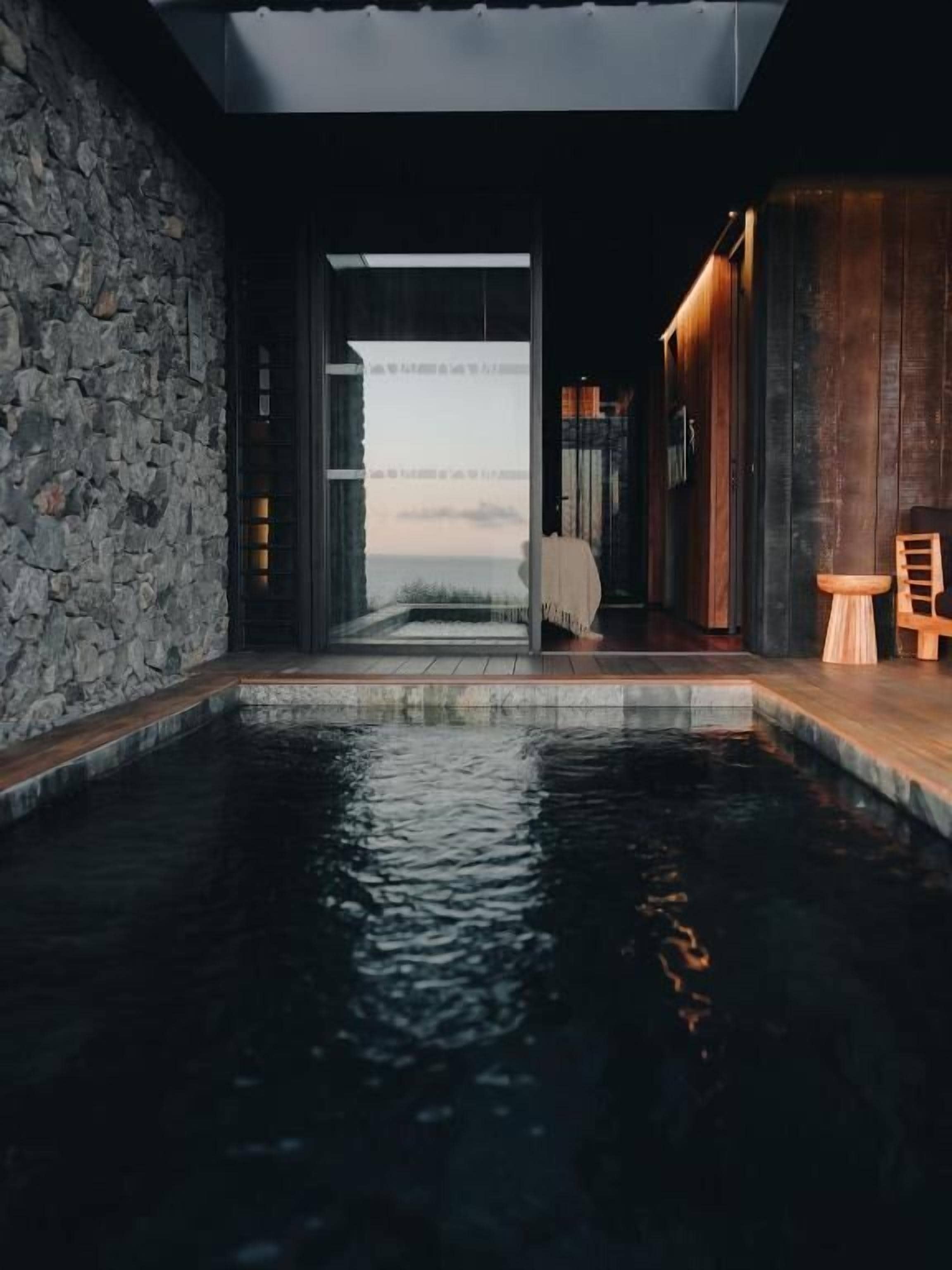 Piscina interior moderna com paredes de pedra, um deck de madeira e vista para o mar.
