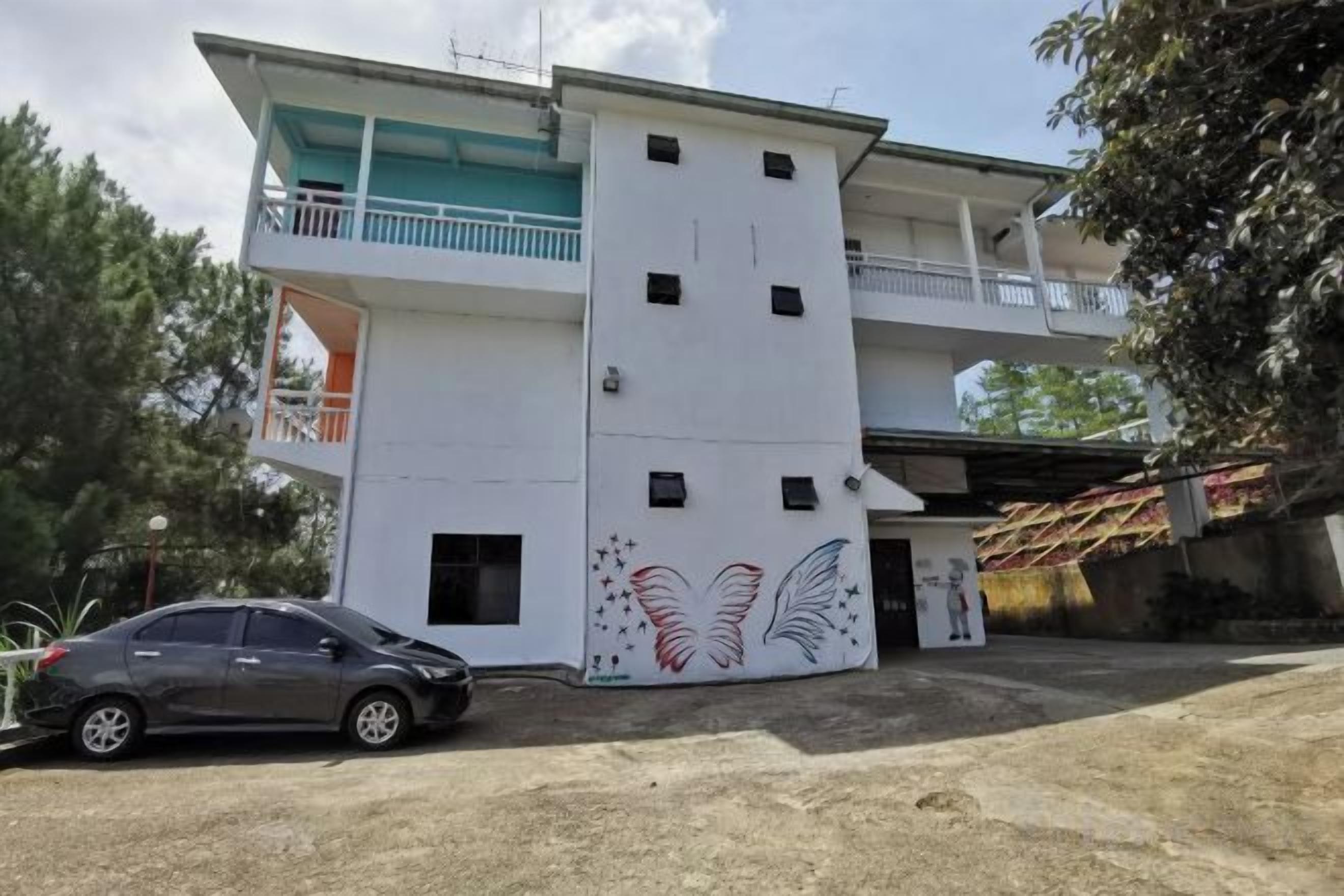 Um edifício de dois andares com varanda no andar superior, um carro estacionado em frente e um muro decorativo com design semelhante a asas.