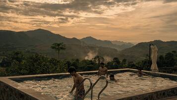 Piscina com três pessoas, cercada por montanhas e vegetação.