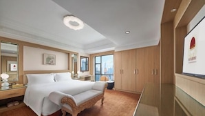 Quarto de hotel com uma cama grande, um guarda-roupa de madeira, uma secretária com uma lâmpada e vista para a cidade pela janela.