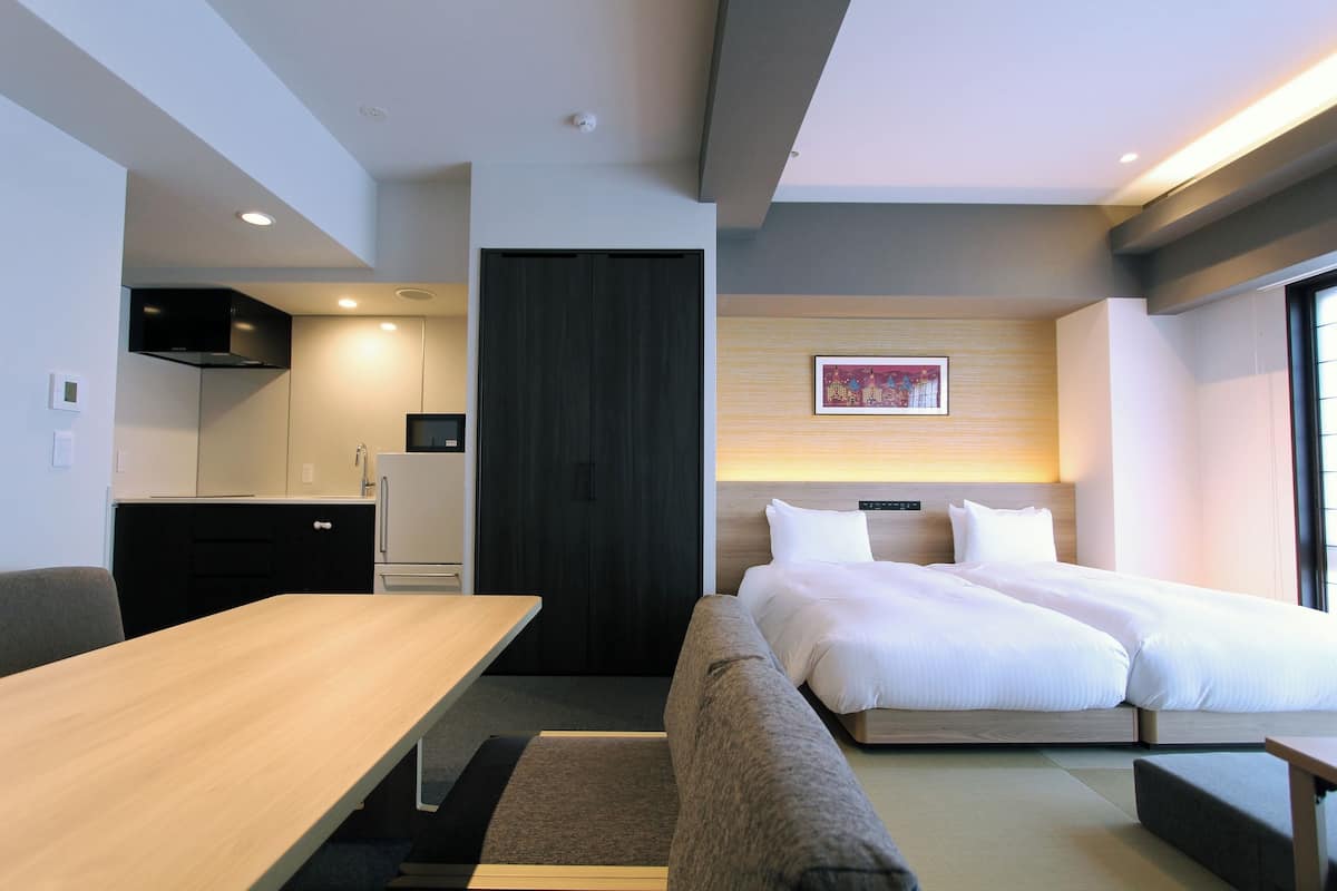 Quarto de hotel moderno com cama, escrivaninha, cadeira, sofá, mesa pequena, televisão e área de cozinha.