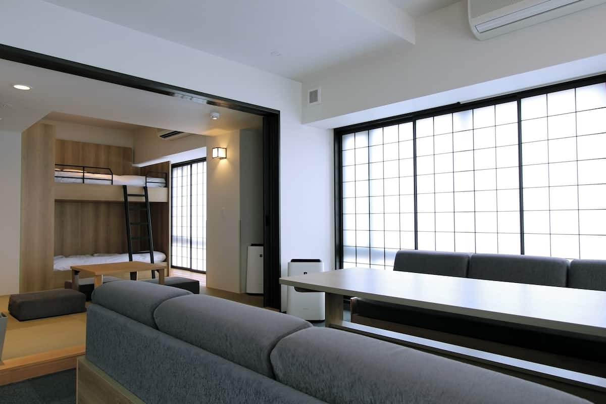 Quarto moderno e compacto com beliche, sofá, mesa de jantar e amplas janelas que proporcionam luz natural.
