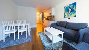 Uma sala de estar moderna com um sofá cinzento, mesa de centro branca e cadeiras brancas. Um quadro está na parede, e uma cozinha é visível ao fundo.