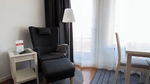 Quarto de hotel moderno com um sofá cinza escuro, uma pequena mesa de apoio branca com um controle remoto, um abajur e uma mesa de jantar com uma cadeira.