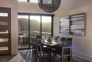Uma sala de jantar com mesa posta para quatro pessoas, uma janela grande com vista para montanhas e duas pinturas abstratas nas paredes.