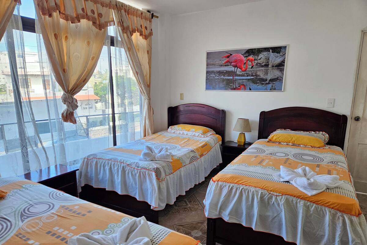 Quarto de hotel com duas camas, uma janela com cortinas, uma mesa de cabeceira com abajur e um quadro de um flamingo.