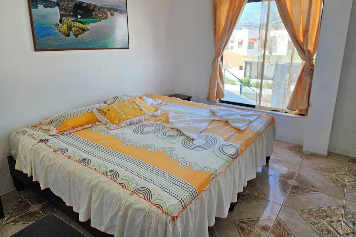 Cama bem arrumada com roupa de cama estampada, janela com cortinas e um quadro na parede.