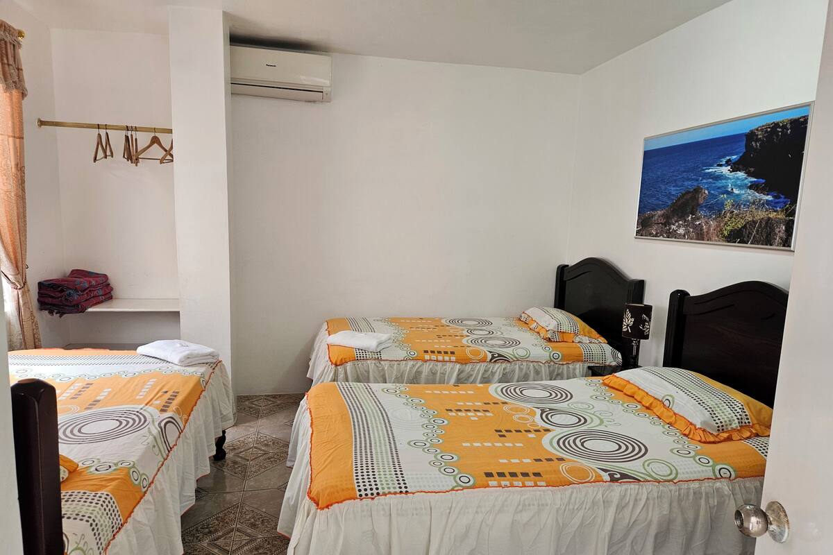 Quarto de hotel com duas camas, um guarda-roupa, um ar condicionado, um quadro de uma paisagem costeira e chão de azulejos.